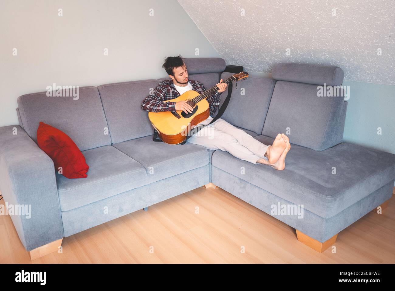 Barefoot musician sits comfortably on a gray couch, strumming an ...