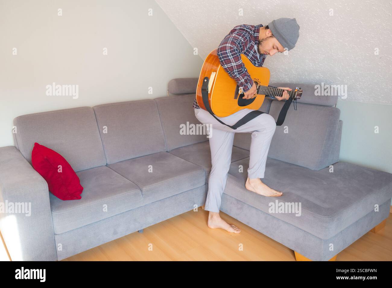 Barefoot musician balances on a gray couch, deeply immersed in playing ...