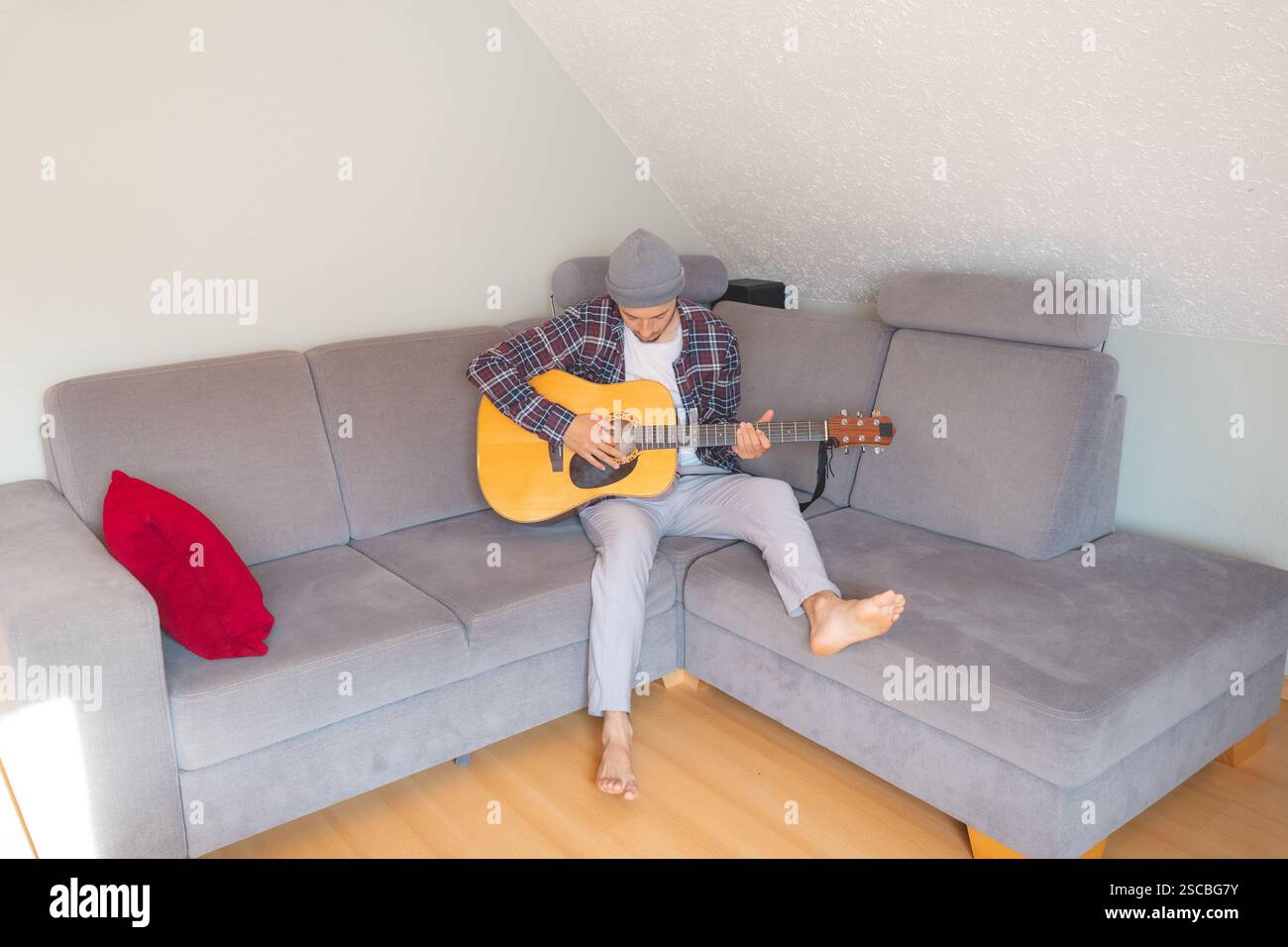 Barefoot musician sits comfortably on a gray couch, strumming an ...