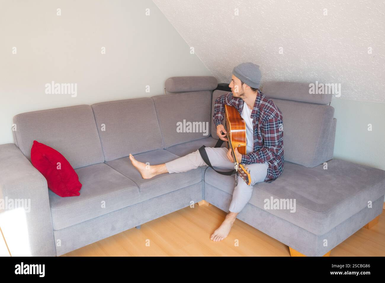 Barefoot musician sits comfortably on a gray couch, strumming an ...