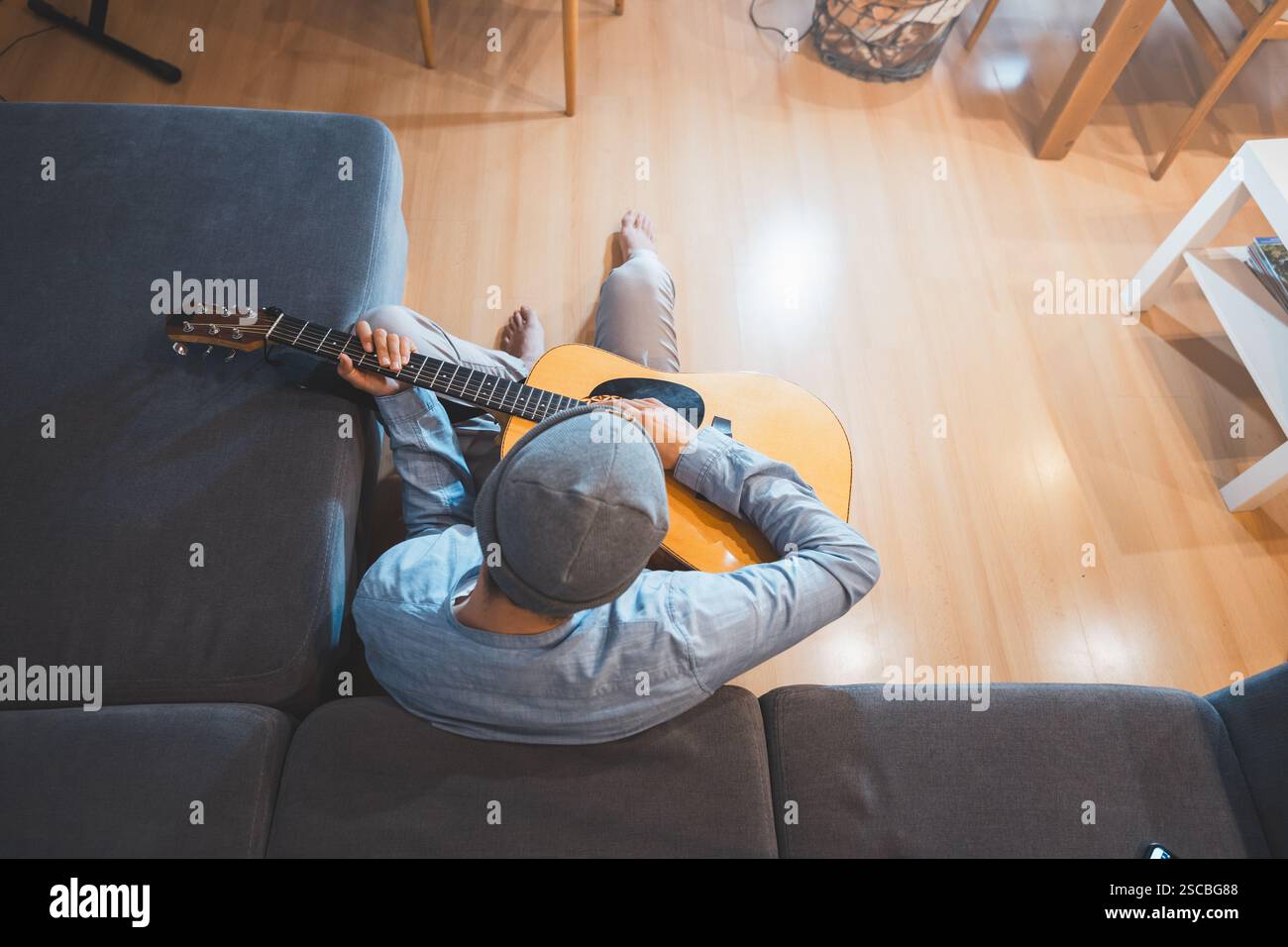 Musician sits on a gray couch, casually strumming an acoustic guitar ...