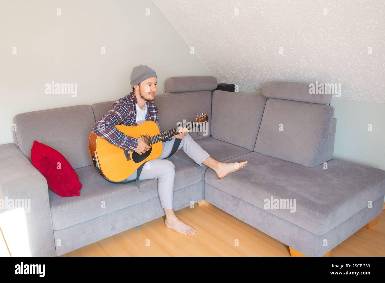 Barefoot musician sits comfortably on a gray couch, strumming an ...