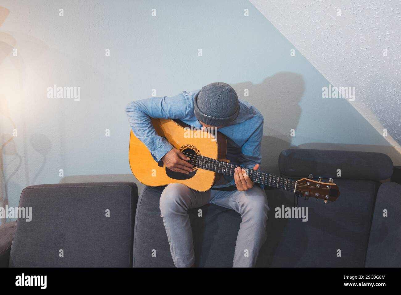 Musician sits on a gray couch, fully absorbed in playing an acoustic ...