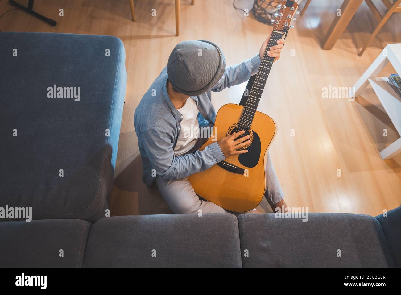 Musician sits on a gray couch, casually strumming an acoustic guitar ...
