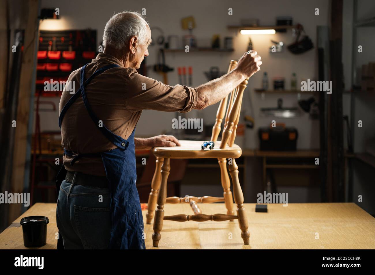 Elderly male carpenter working with old chair for restoration in dark ...