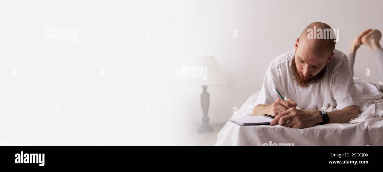 Millennial man taking notes in his diary while relaxing on bed at home ...