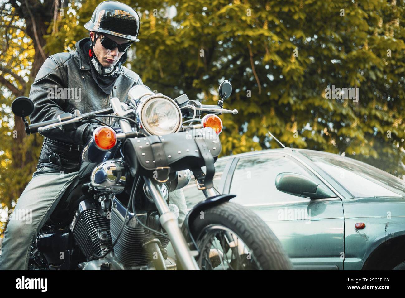Biker on a classic motorcycle next to a parked car under trees Stock ...