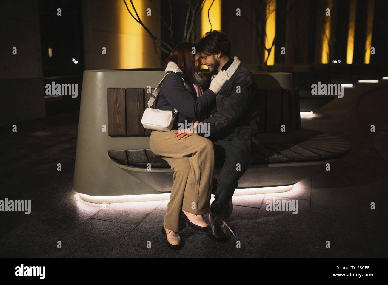 Couple sitting closely on a bench at night in a city, sharing an ...