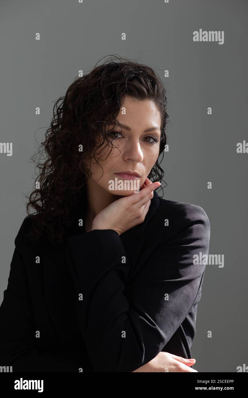Thoughtful woman with curly hair in a black suit, resting her chin on ...