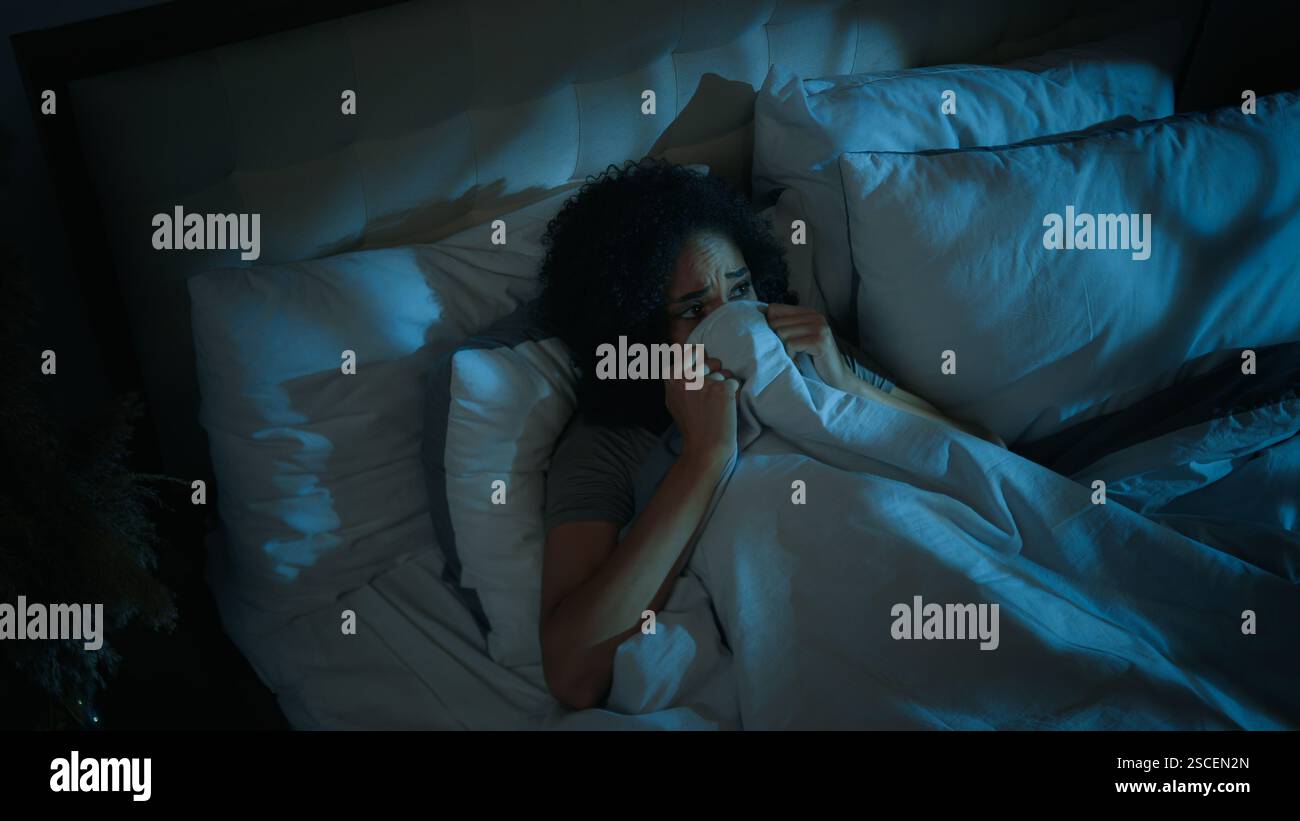 Scared frightened African American woman hiding under blanket bed nightmare sleep disorder bad ...