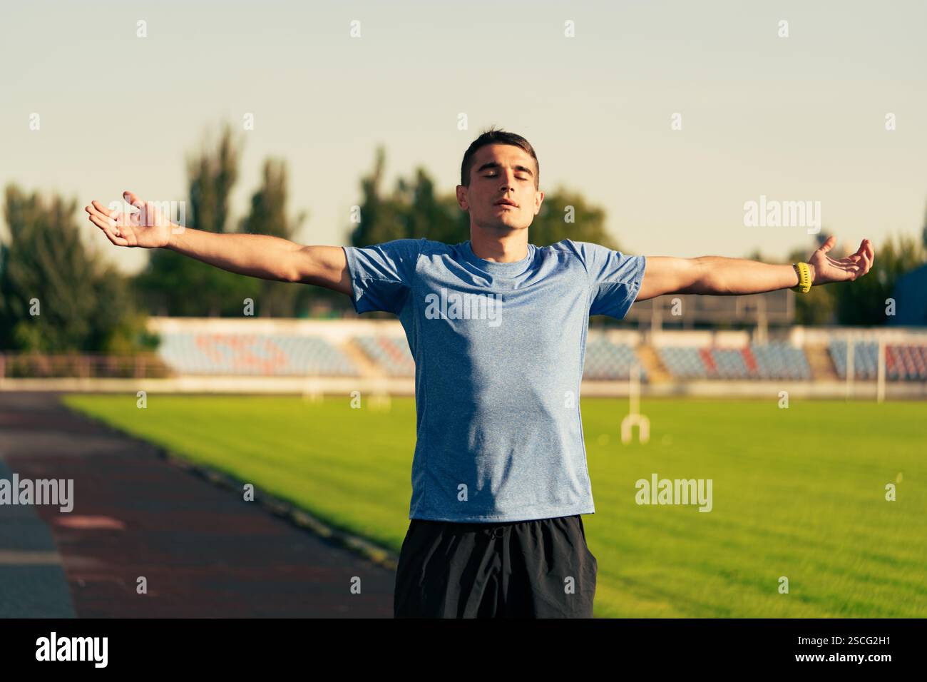 Young man stretches with arms wide open on a track field during sunset ...