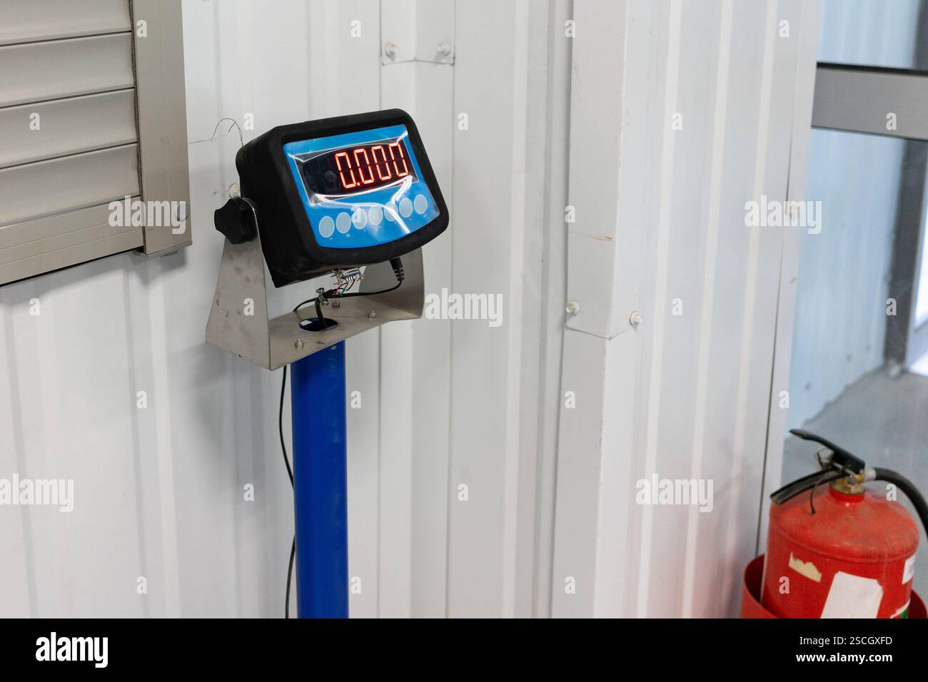 A digital scale stands against a white wall, displaying a weight of ...