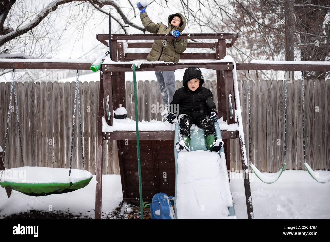 Two kids enjoy snowy fun on a backyard wooden playset and slide Stock ...