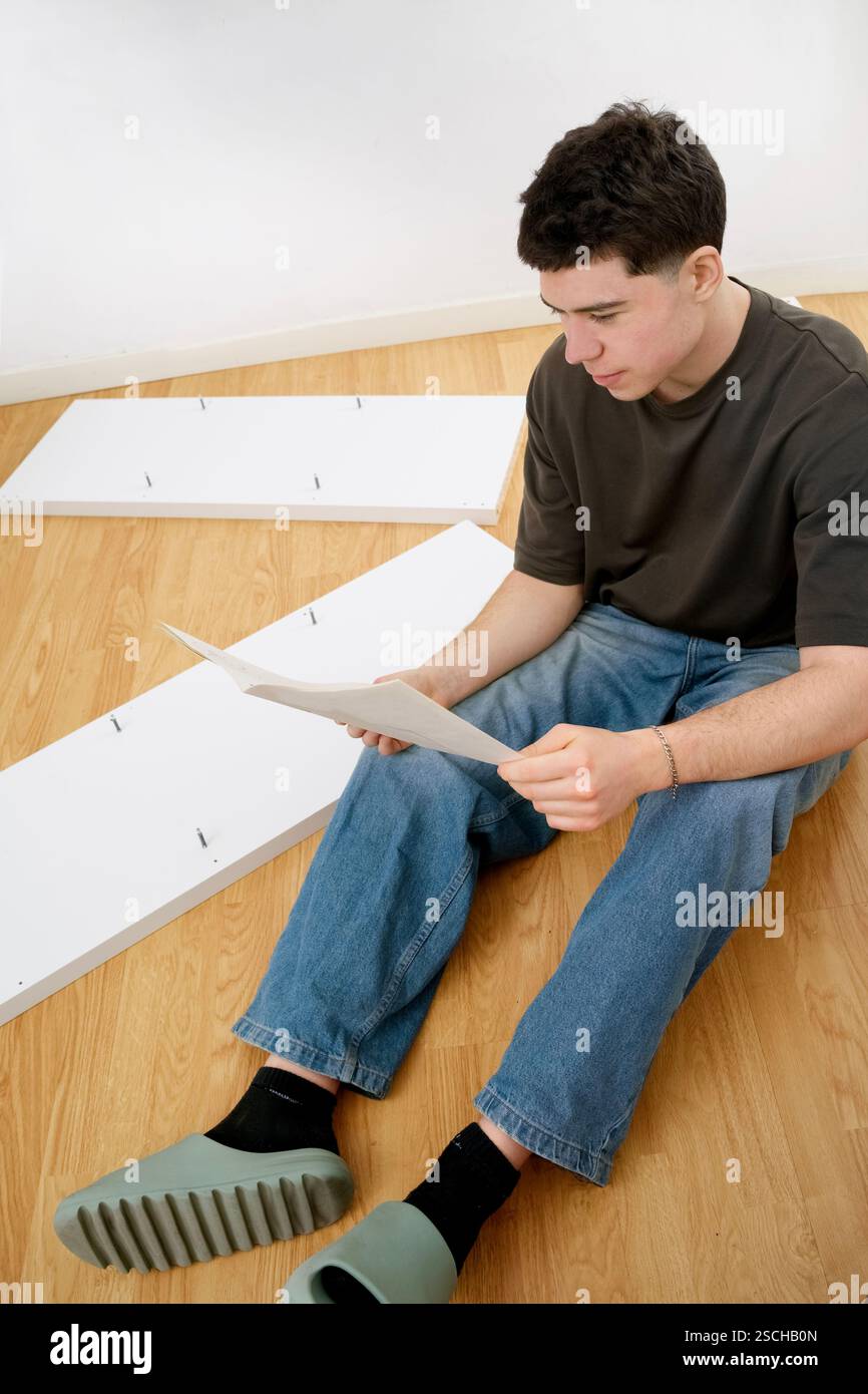 A young man sits on a wooden floor, reading assembly instructions for ...