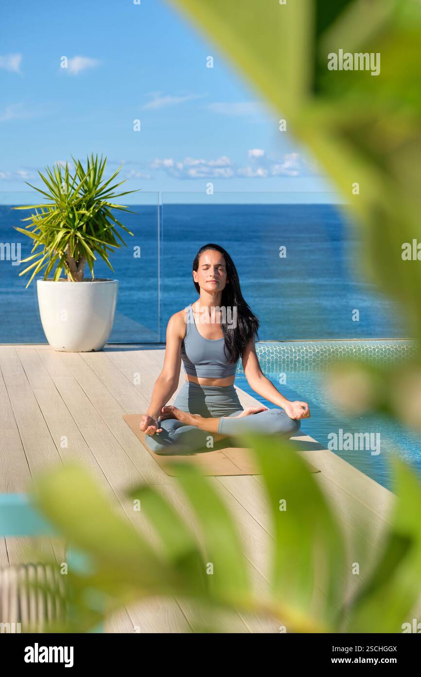 A woman practices meditation on a deck overlooking the ocean. She sits cross legged, eyes closed ...