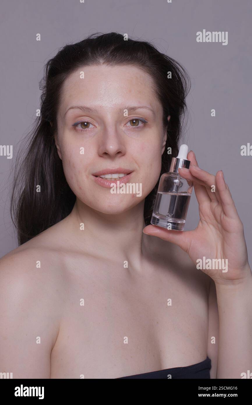 Woman without makeup with glowing skin holding a bottle of clear serum ...