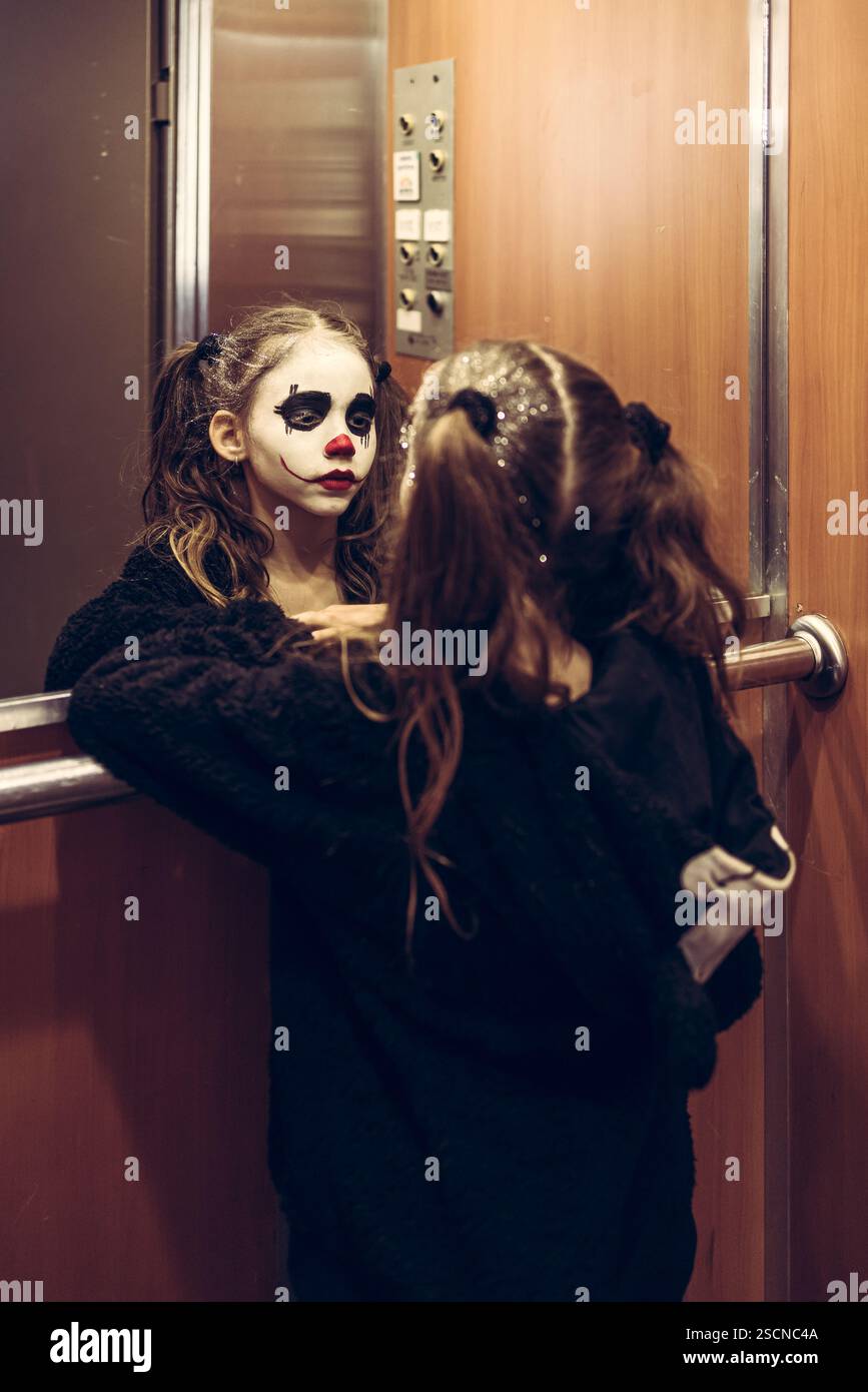 Beautiful 9-year-old gothic girl with sad clown makeup watching herself in the mirror inside the ...