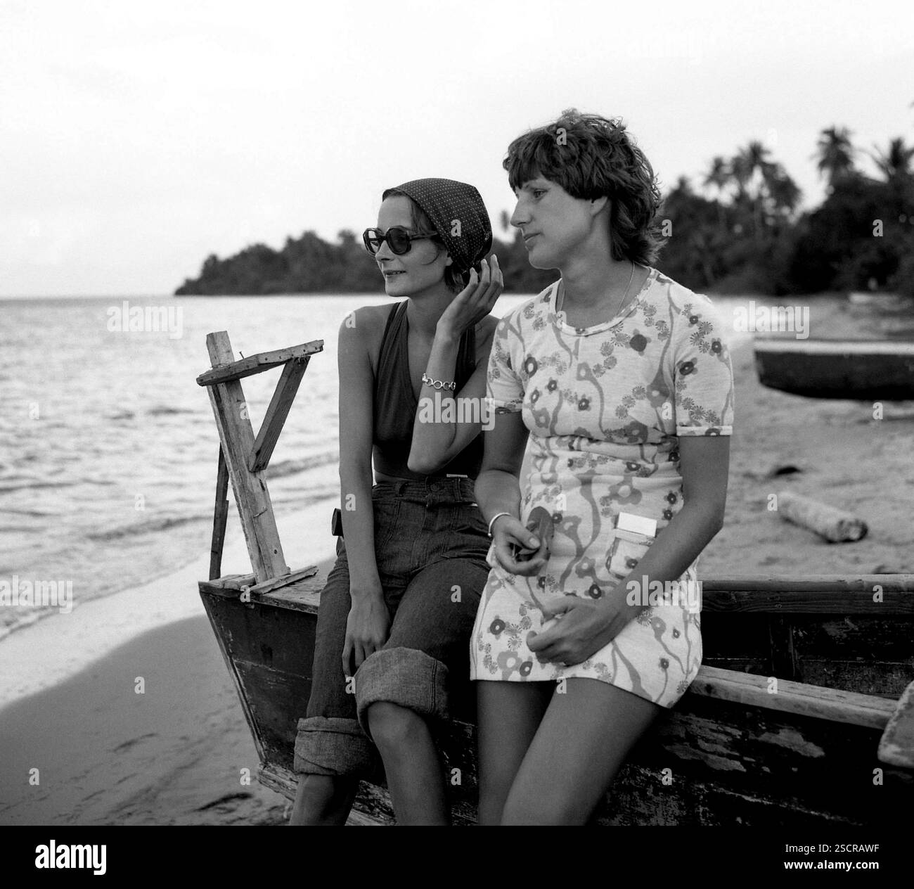 Two women lean against a rowing boat on a beach in Jamaica and look out ...