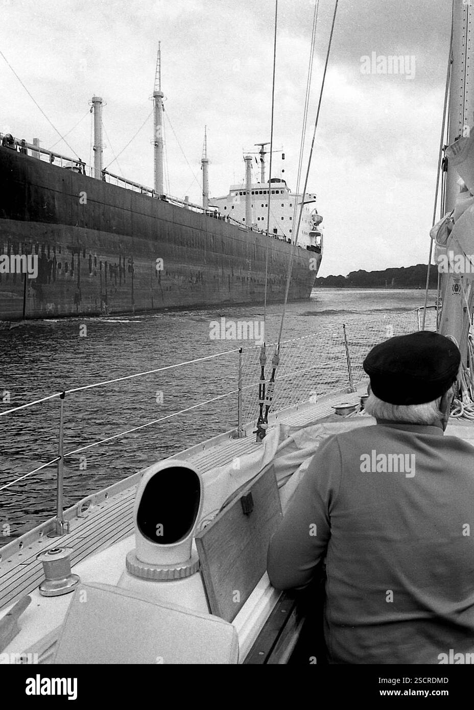 An elderly man steers a sailing boat past a much larger cargo ship in ...