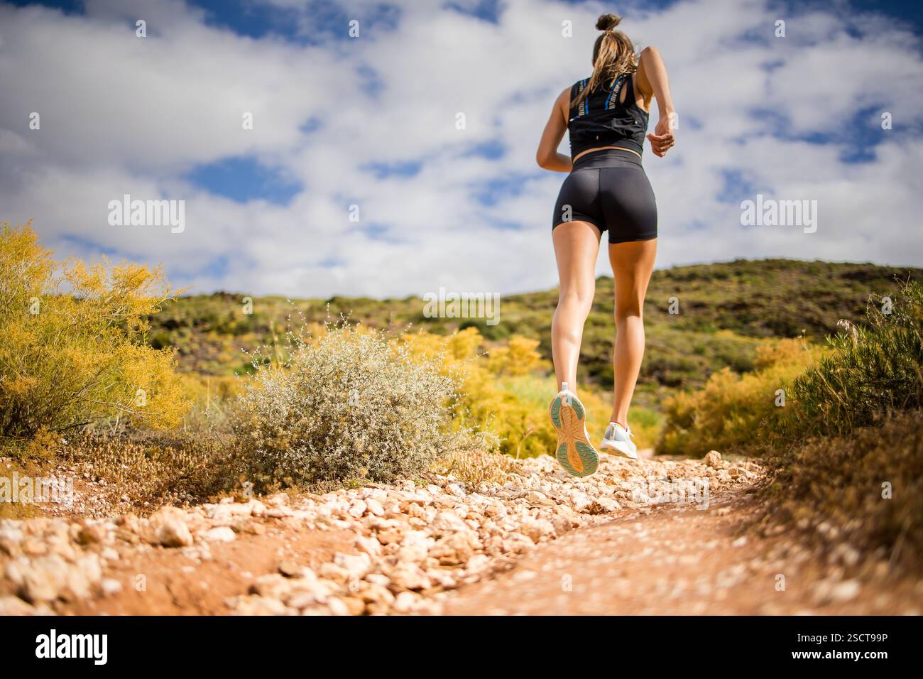 Female trail runner wearing black sportswear and a hydration vest, running through a scenic ...