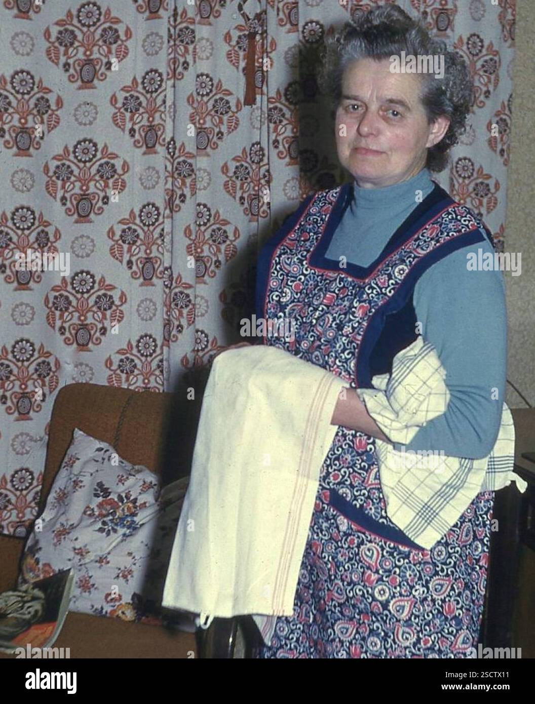 An elderly woman wearing a Dederon apron stands in front of a patterned ...
