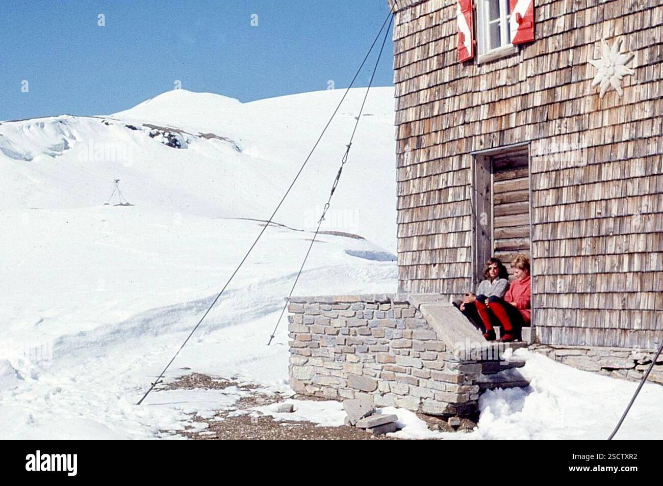 Two women sunbathe sheltered from the wind in front of a hut in the Alps. [automated translation ...