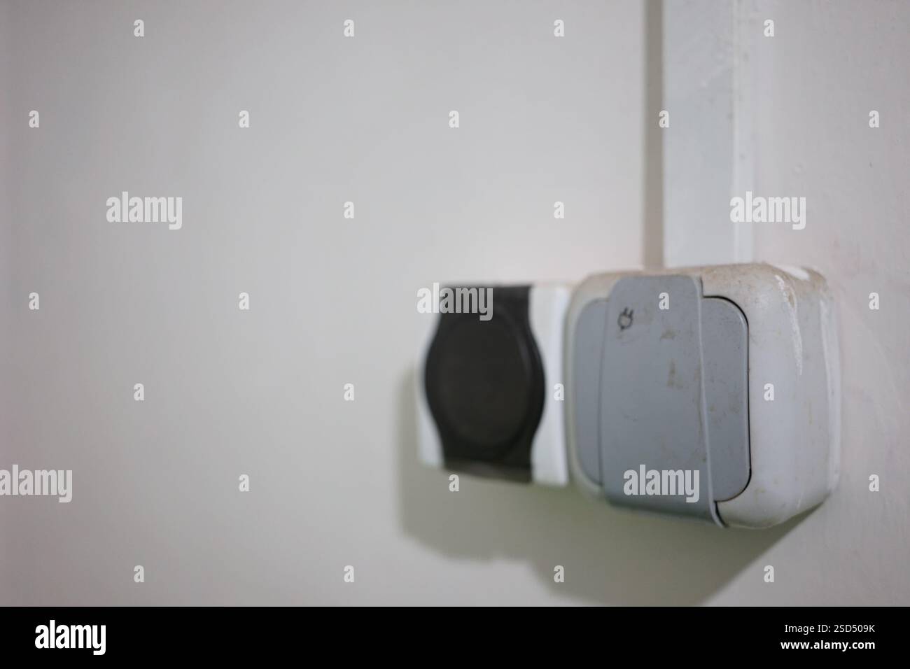 Two electrical switches are fixed to a plain white wall in a room. One ...