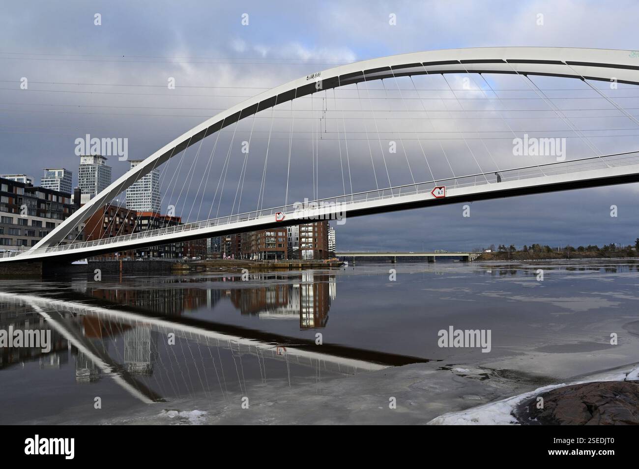 A modern white arch pedestrian bridge in Helsinki, Finland, spans over a calm river, reflecting ...