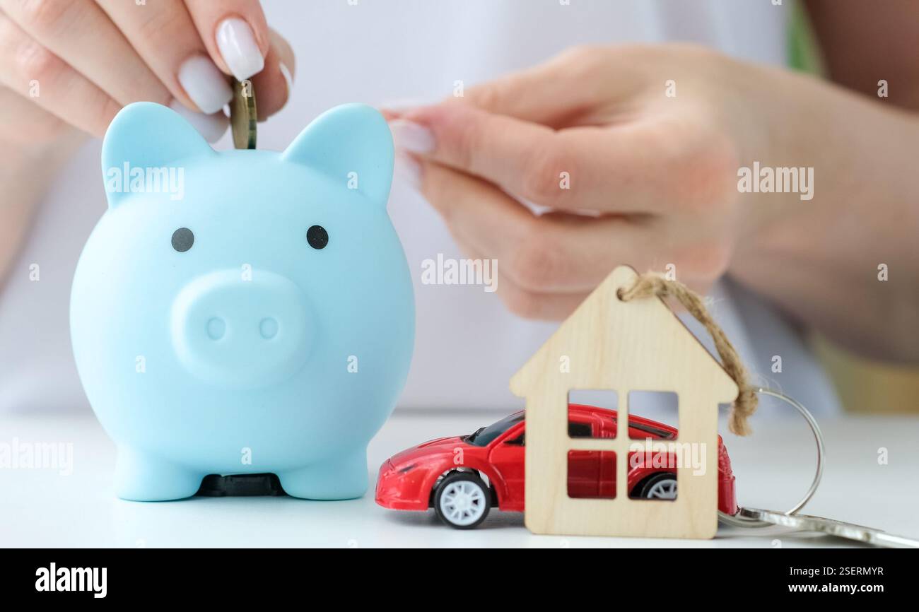 Hand putting coin into blue piggy bank next to house key and red toy ...