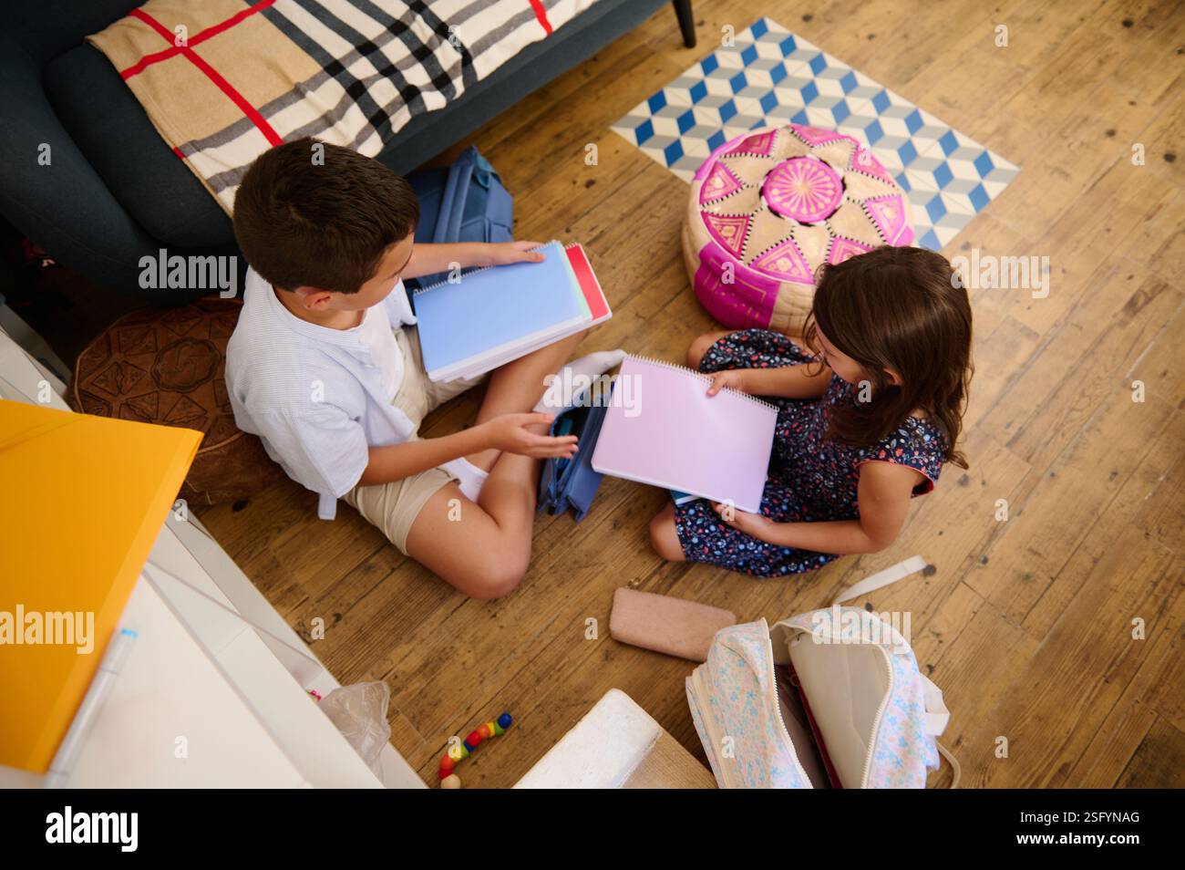 Two children sit on a wooden floor organizing their school supplies ...