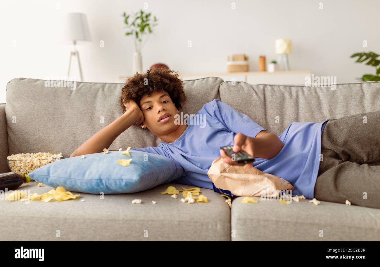 Lazy black teen guy lying on sofa with scattered food, holding TV ...