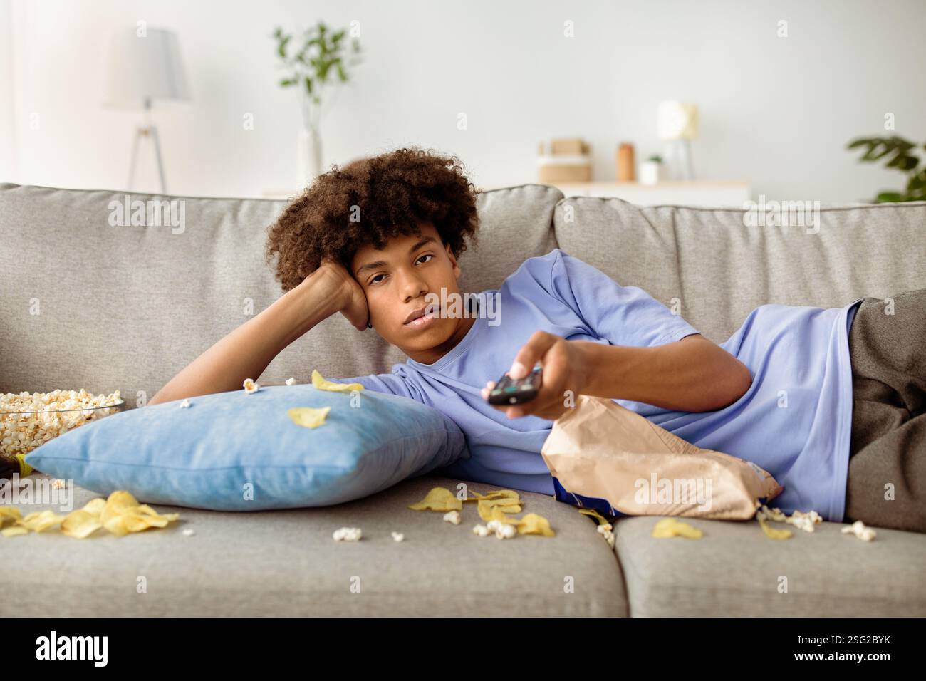 Lazy black teen guy lying on couch with scattered snacks, holding ...