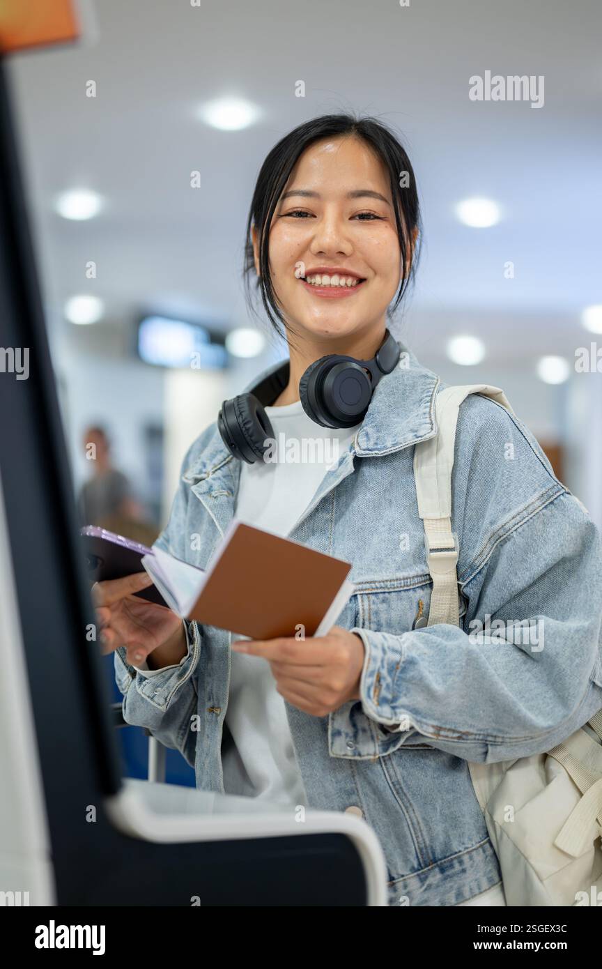 A beautiful, smiling Asian female passenger or traveler stands in front of an airport self-check ...