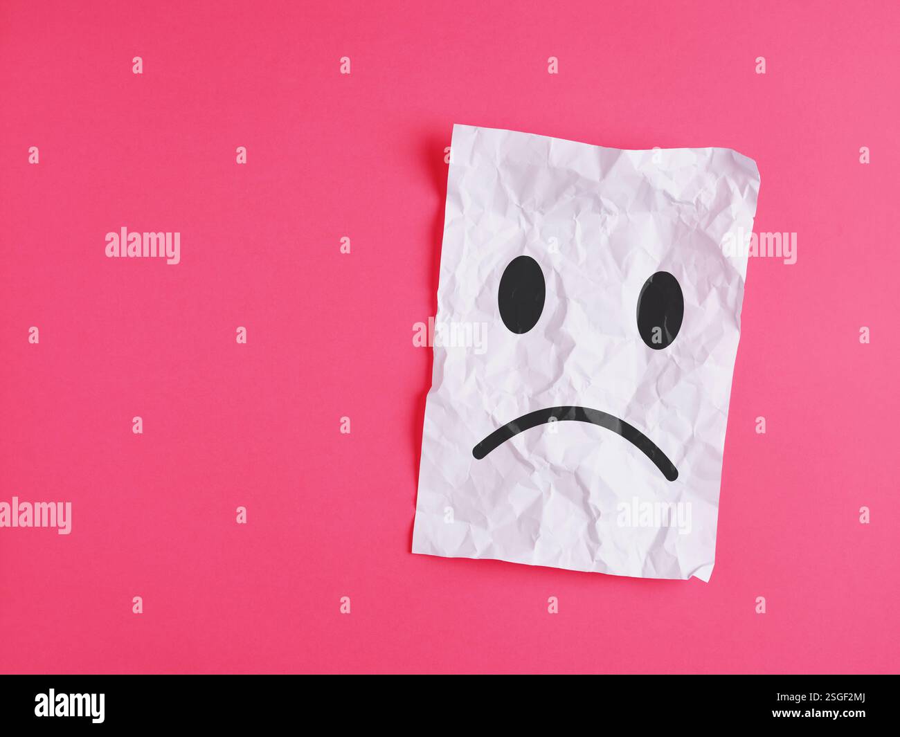 Sad face drawn on crumpled paper is lying on pink background, conveying ...
