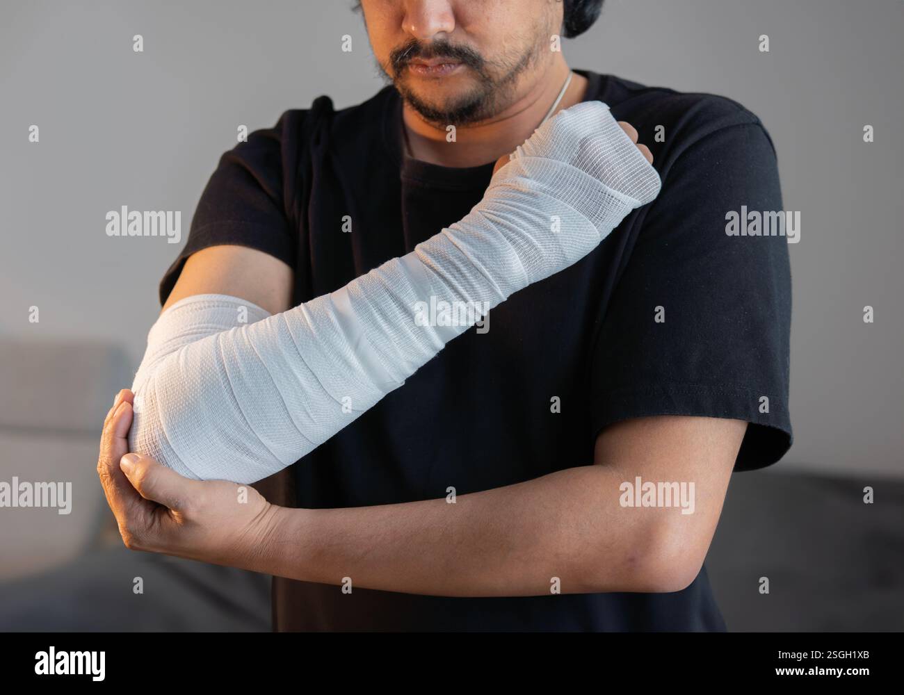 Beard man with arm wrapped in medical bandage sit on sofa over light ...