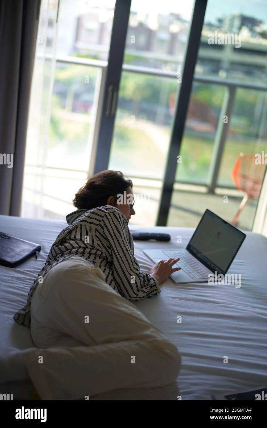 Woman lying on a bed working on a laptop in a bright room with large ...