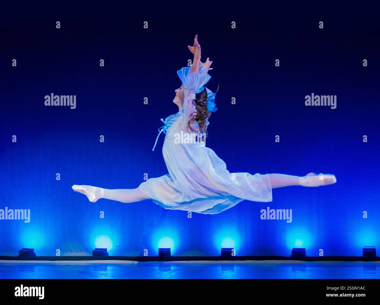 Ballerina in a flowing dress performs a split leap on stage with blue ...