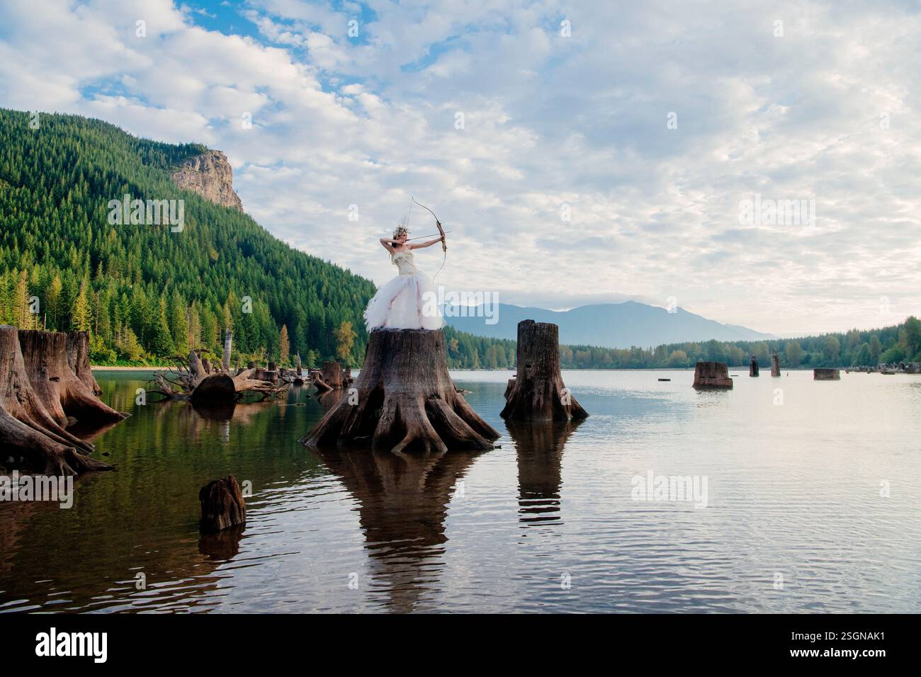 Woman in a white dress aiming a bow from a tree stump in a serene lake ...