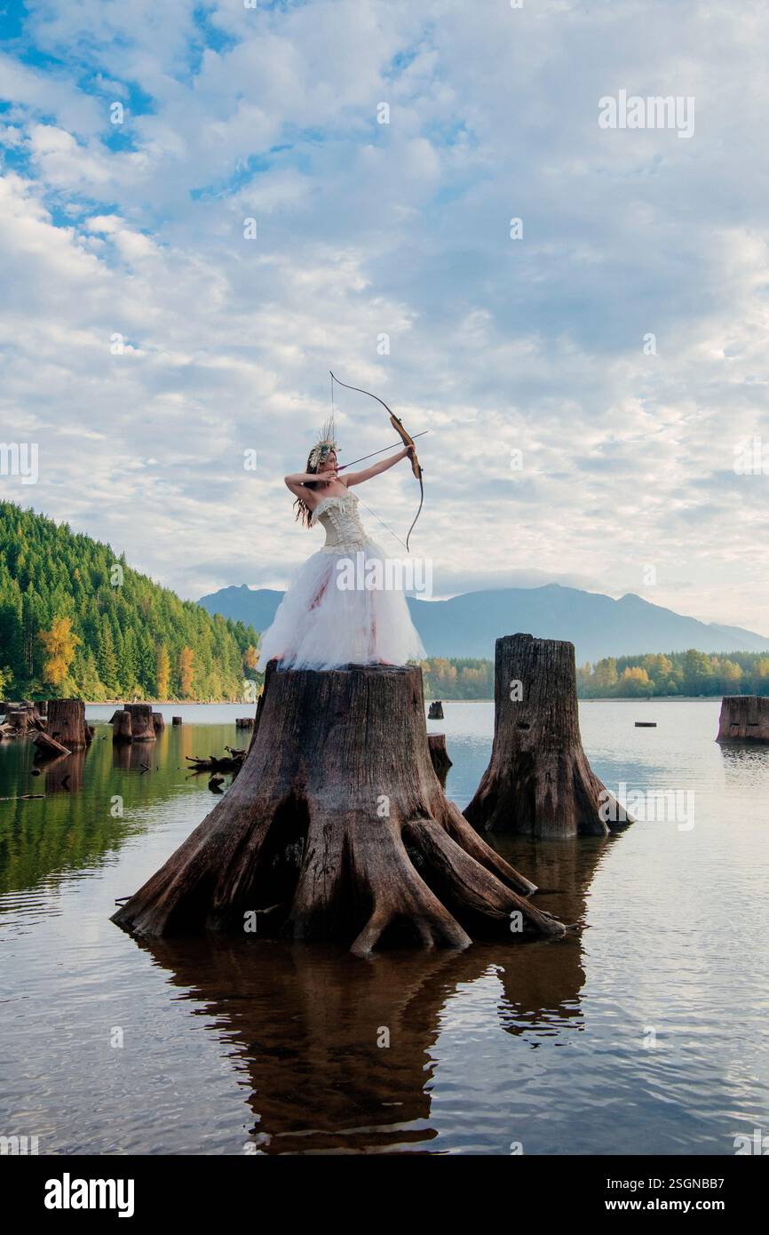 Woman in a white dress with a bow and arrow standing on a large tree ...