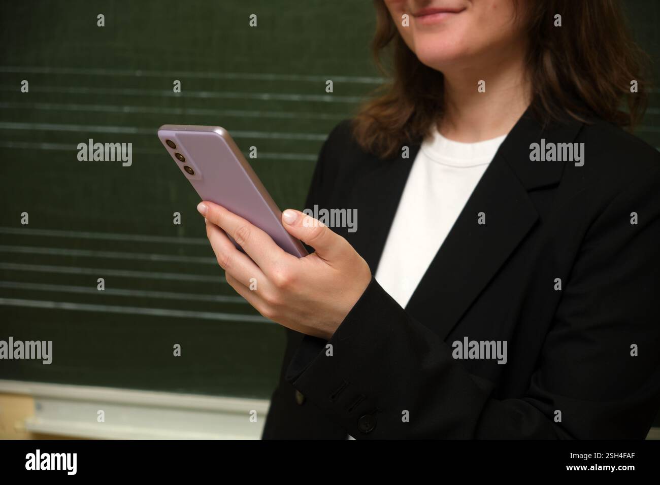 The female teacher, standing by the blackboard in her jacket, is holding a smartphone, showing ...