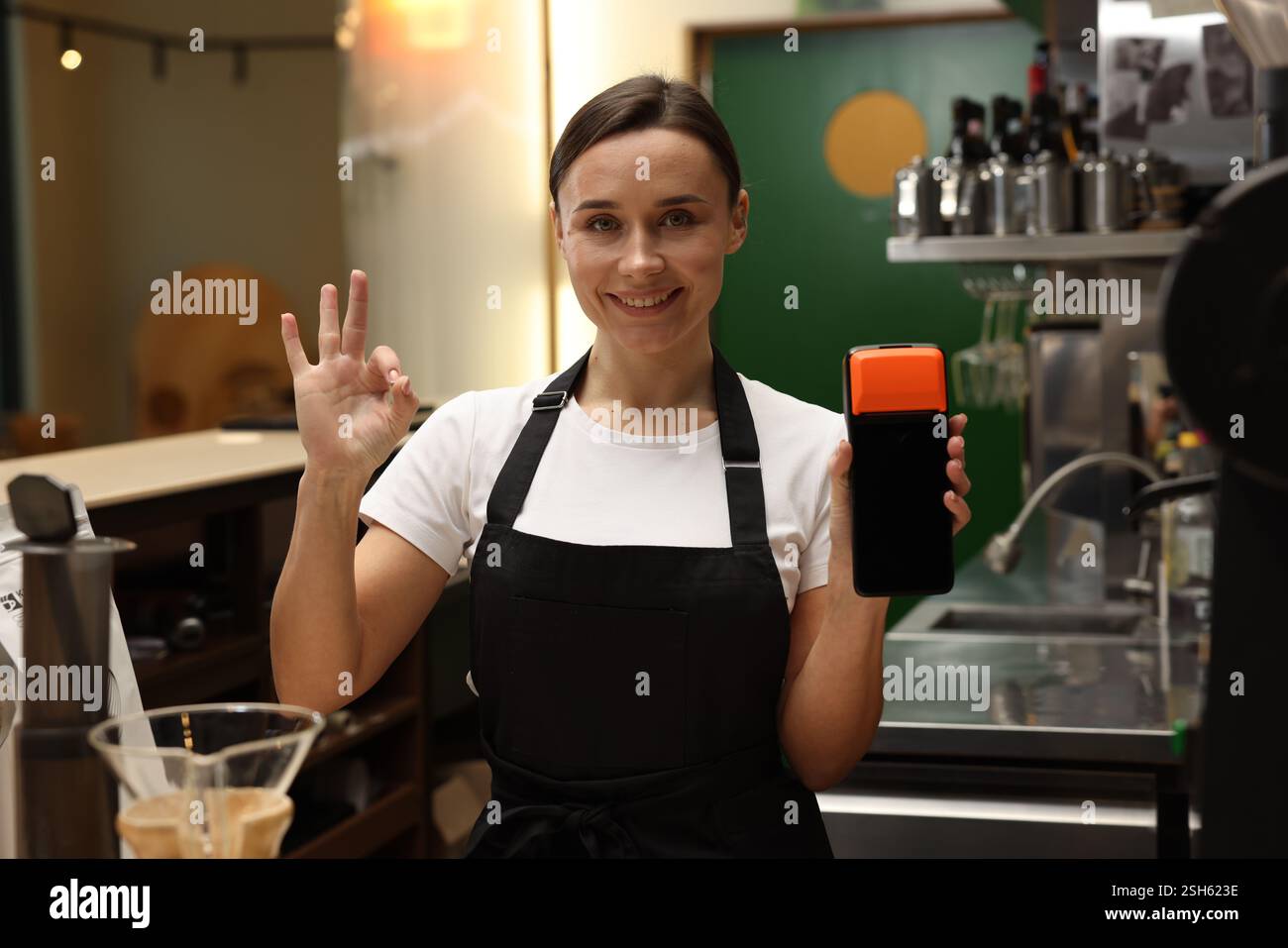 Smiling cafe worker with payment terminal showing OK gesture indoors ...