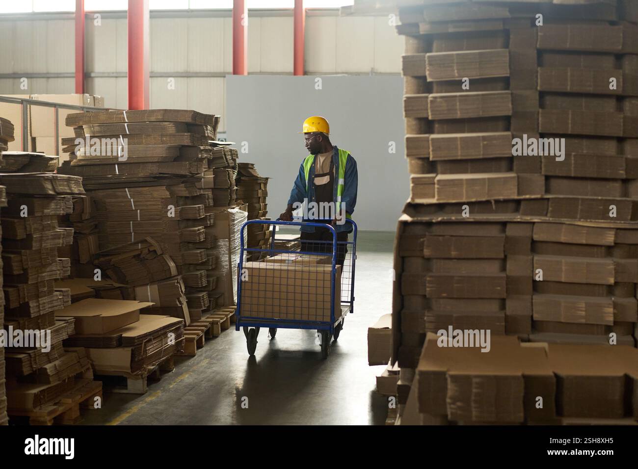 Young male engineer pushing cart with packed boxes while moving between stacks of cardboard ...