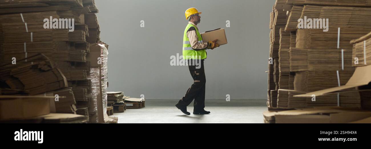 Header of male engineer carrying packed box on hands while moving between tall stacks of ...