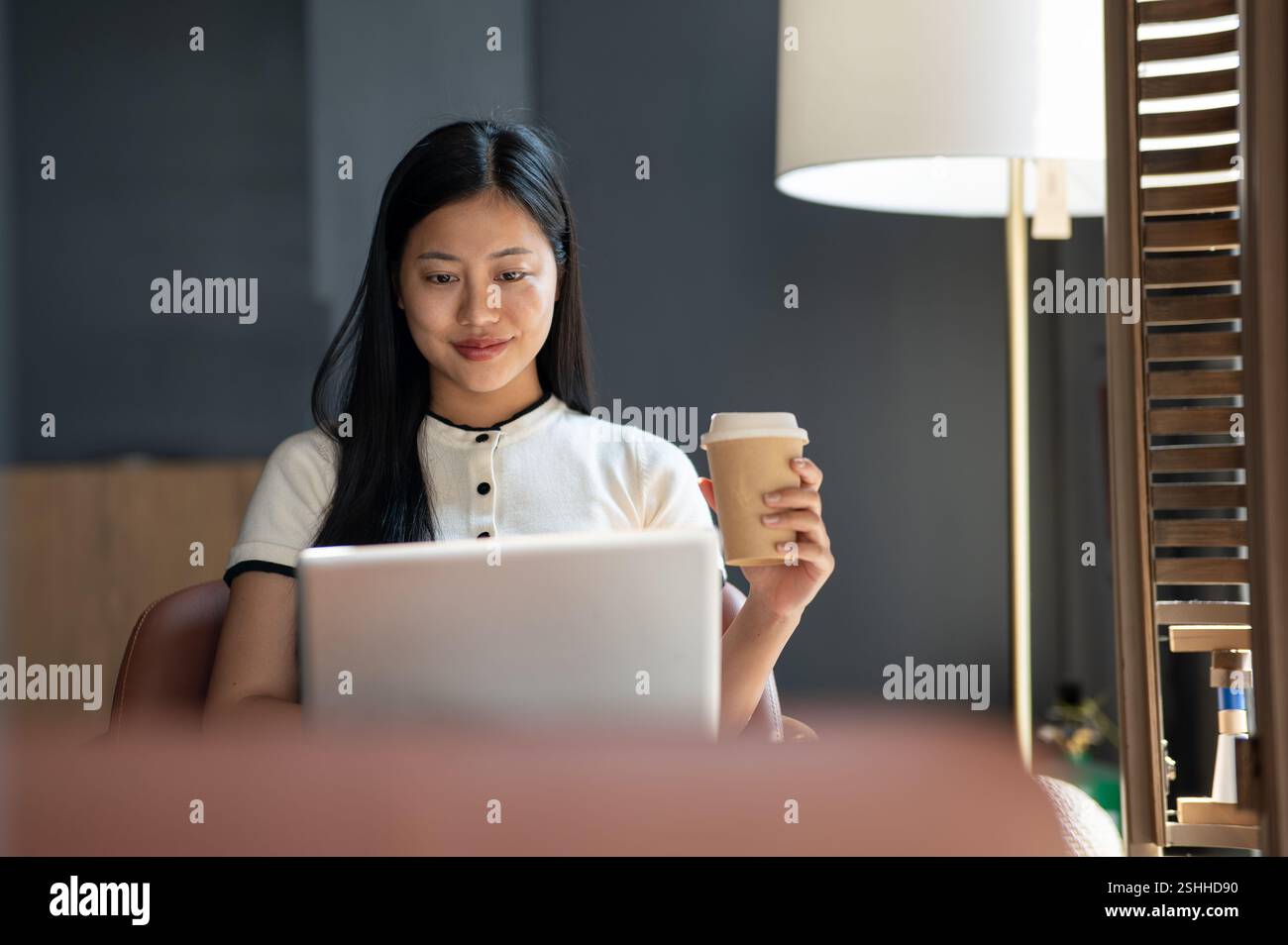 A positive Asian woman works remotely from a coffee shop, carefully ...