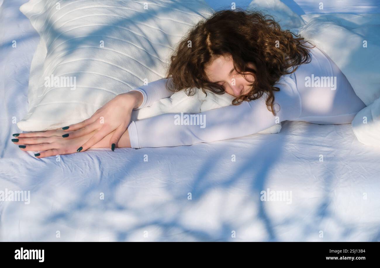 Young woman lies on white sheets, her face partly hidden in the pillow ...