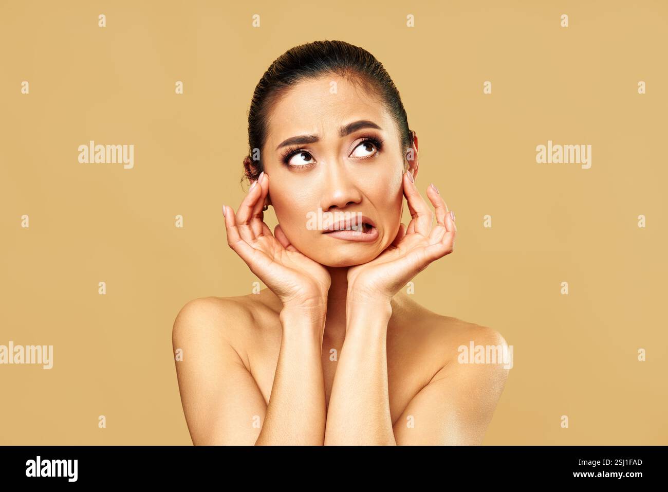 Worried young woman biting her lip, looking up over beige background ...