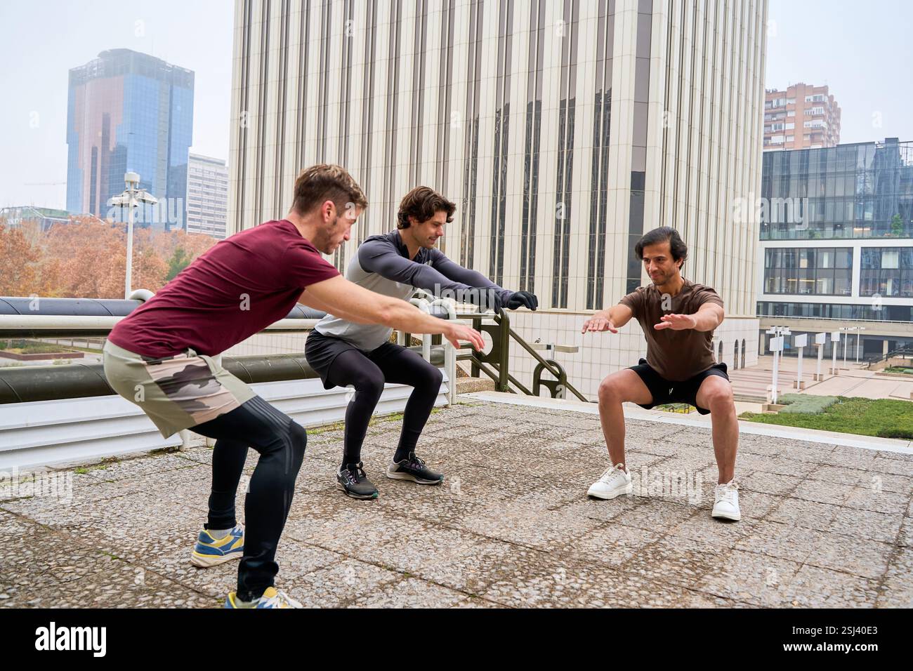 Three male athletes performing squats exercises outdoors in an urban ...