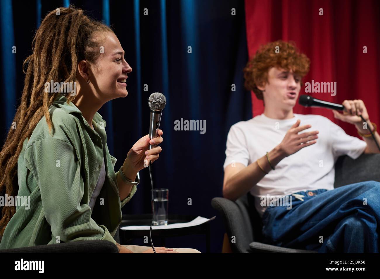 Two young adults seated in chairs laughing while holding microphones ...