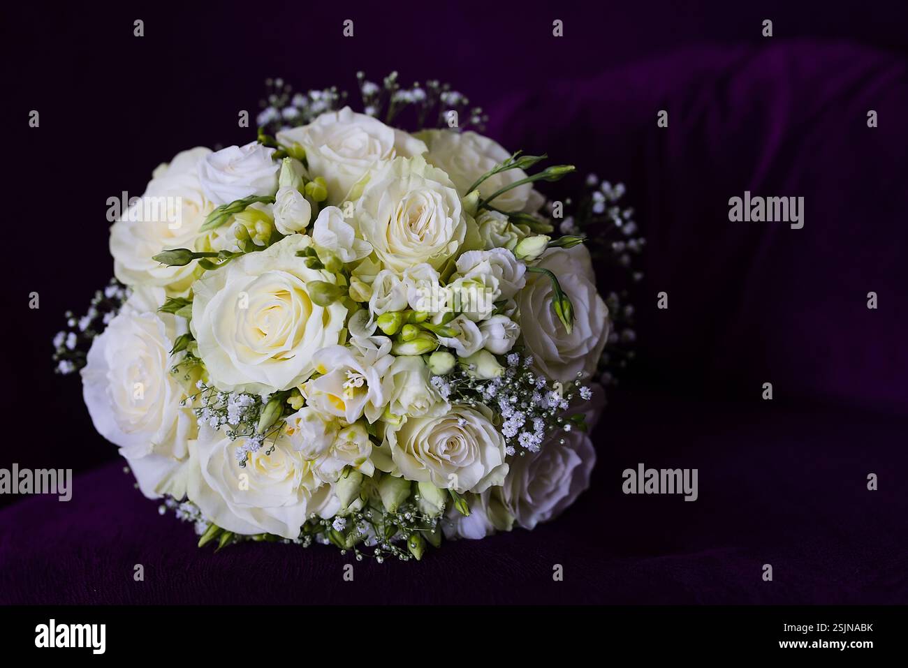 A stunning wedding bouquet of white roses, lisianthus, and baby's breath rests against a dark ...