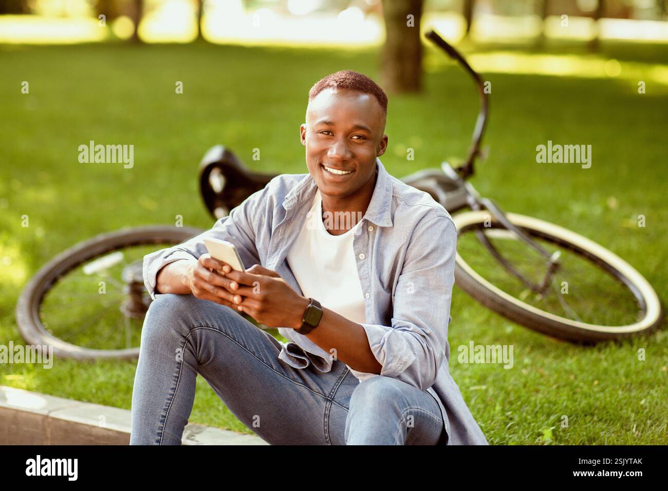 Smiling black guy with mobile phone sitting near his bicycle at park ...